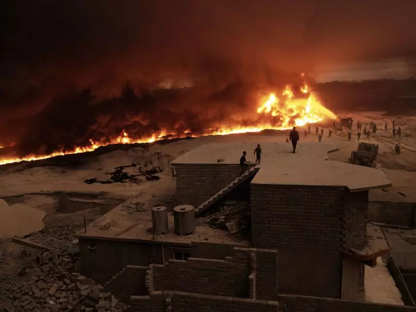 Intsapho ijonge eluphahleni lwayo njengoko abacimi-mlilo bezama ukucima udonga lwamadangatye asondela kufutshane nekhaya labo. Qayyarah, iNiniva Governorate, Iraq, October 26, 2016. From [We came from Fire ]( https://amzn.to/2L9l8Vm) nguJoey L. – epapashwe yi powerHouse Books