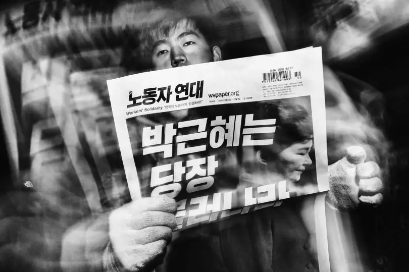 Lês der alles oer. Tidens in kâlde winterjûn ferkeapet in man arbeiderssolidariteitskranten oan demonstranten dy't opmarsjeare nei it presidinsjele Blauwe Hûs. De kop lêst: "Park Geun-hye Resign Now." © Argus Paul Estabrook. Street Series Winner, Magnum en LensCulture Photography Awards 2017