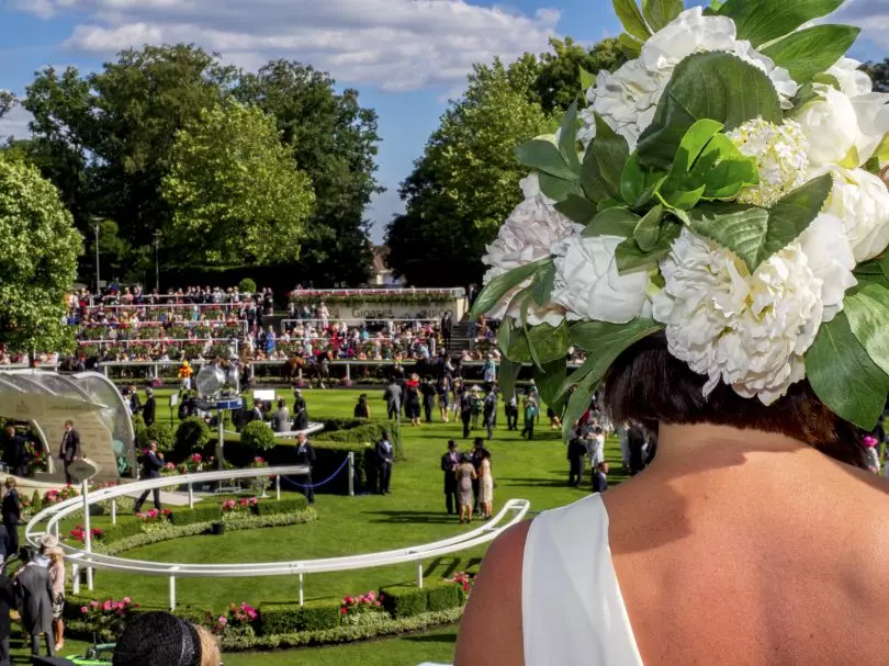 O femeie cu o pălărie decorată extravagant privește peste inelul paradei de Ziua Doamnelor la Royal Ascot. Iunie 2018 © Peter Dench